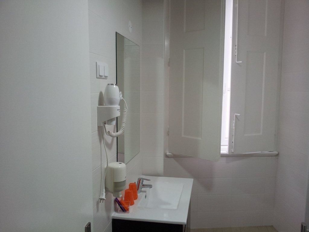 Casa de banho com uma pia branca, espelho e secador de cabelo numa prateleira fixada na parede.