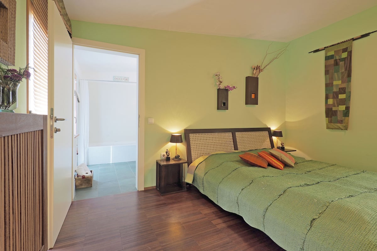 Quarto com colcha verde, piso de madeira, mesa de cabeceira com abajur e porta que dá para outro cômodo.