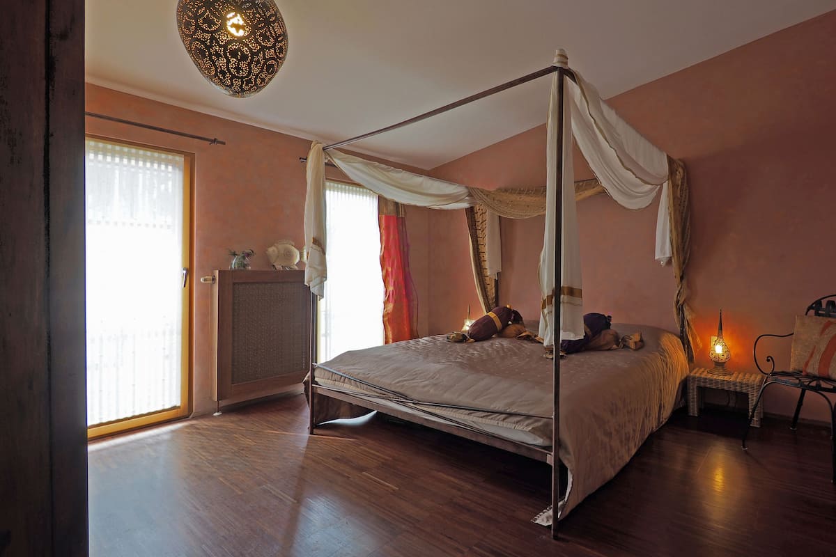 Uma cama com dossel e cortinas, um criado-mudo com luminária, uma cadeira e um guarda-roupa.
