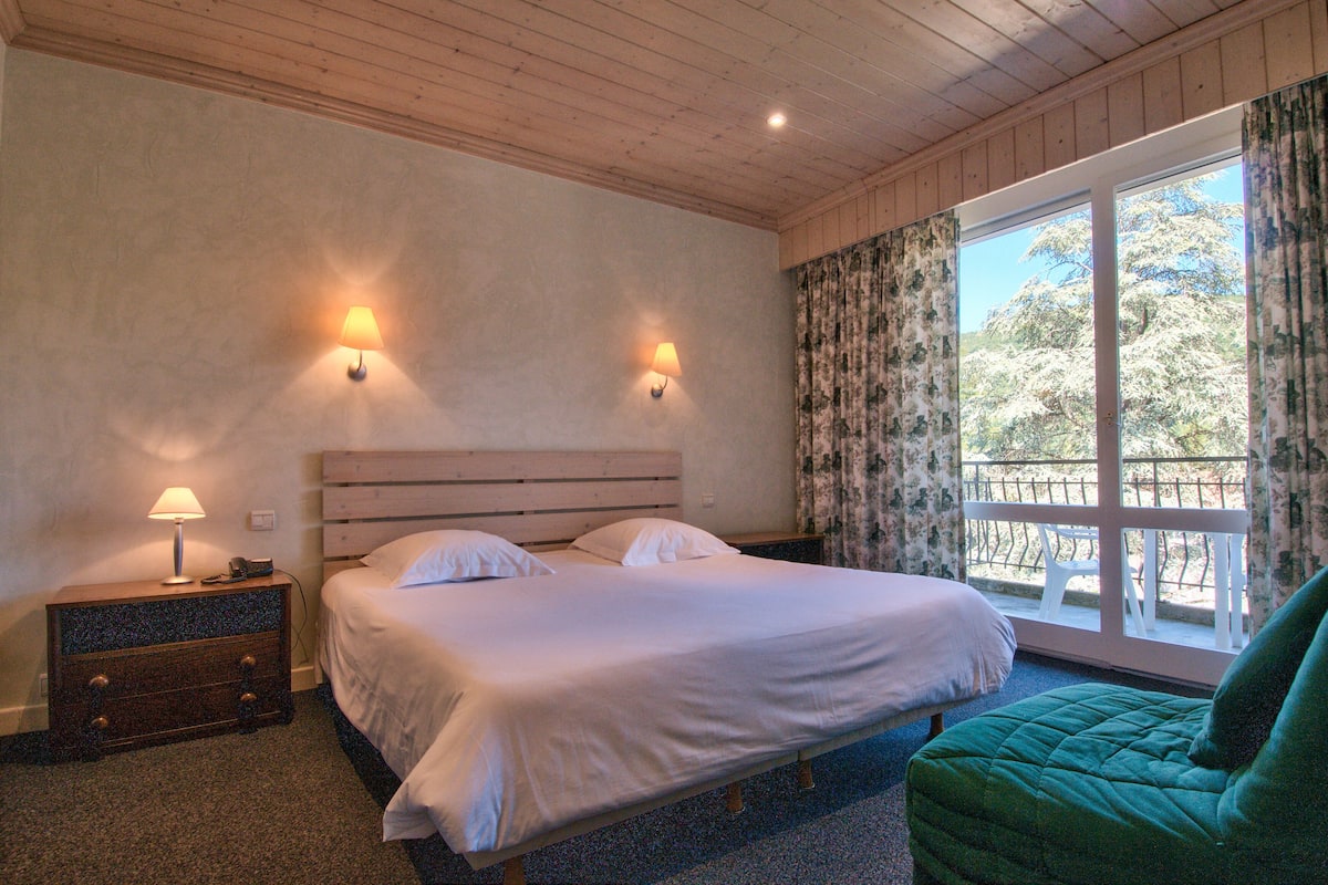 Quarto com uma cama grande, duas luminárias de cabeceira, uma cabeceira de madeira e vista para árvores através de uma janela com cortinas.