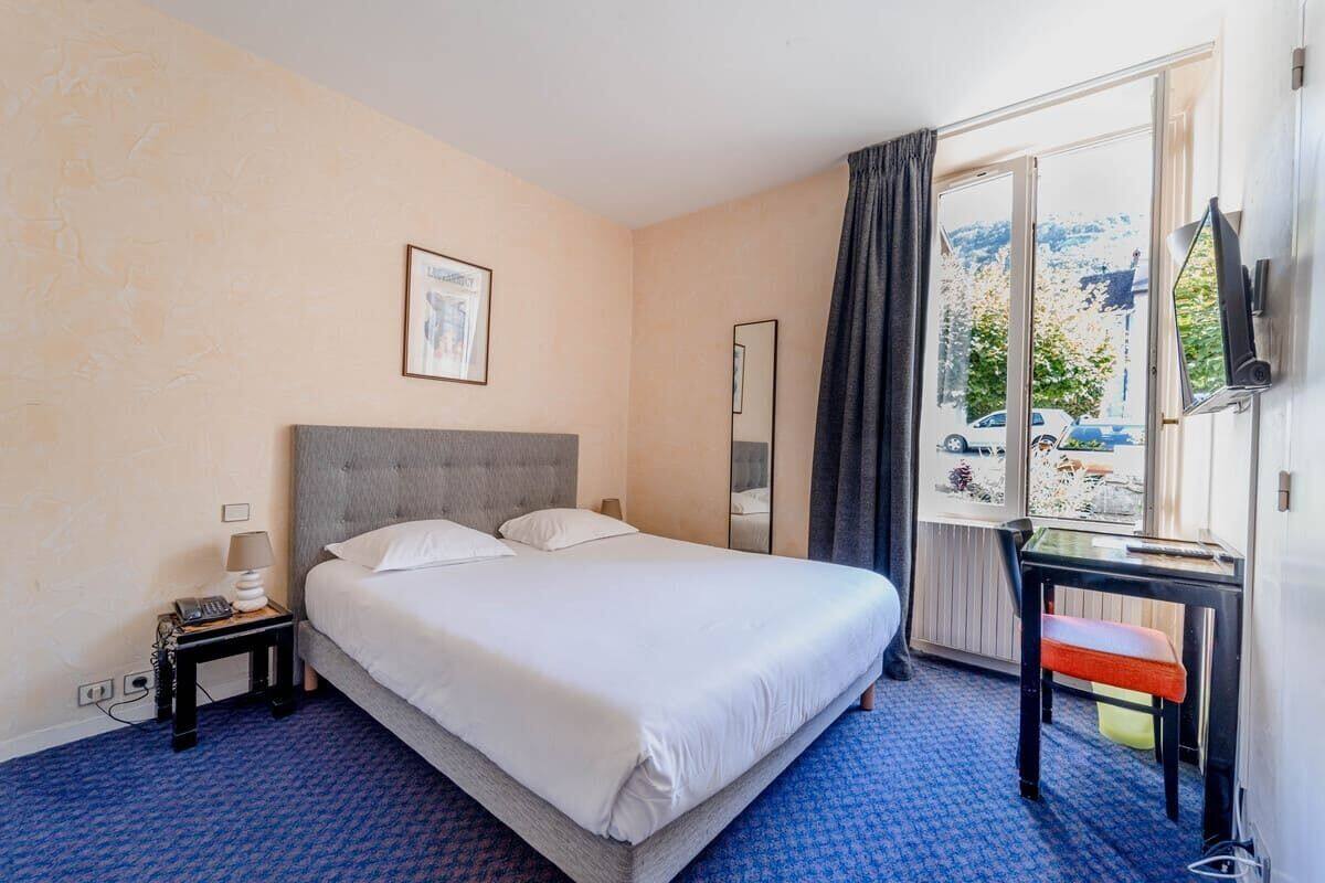 Quarto de hotel com uma cama, mesa de cabeceira com abajur, espelho, televisão e janela com vista para o exterior.