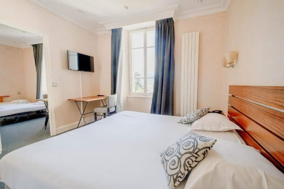 Quarto de hotel com uma cama grande, uma escrivaninha, uma cadeira, televisão e janela com cortinas.