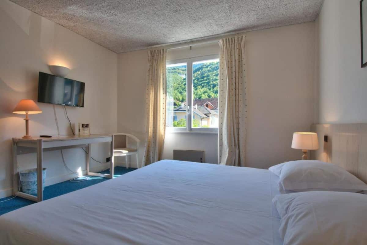 Quarto de hotel com cama, escrivaninha com cadeira, televisão fixada na parede e janela com cortinas.