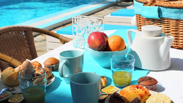 Disposição para o café da manhã perto de uma piscina, incluindo uma cadeira de vime, uma mesa com toalha azul, jarra branca, caneca azul, copo de suco, muffins e uma tigela de frutas.