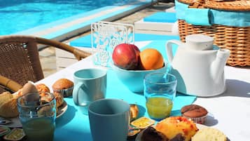 Disposição para o café da manhã perto de uma piscina, incluindo uma cadeira de vime, uma mesa com toalha azul, jarra branca, caneca azul, copo de suco, muffins e uma tigela de frutas.