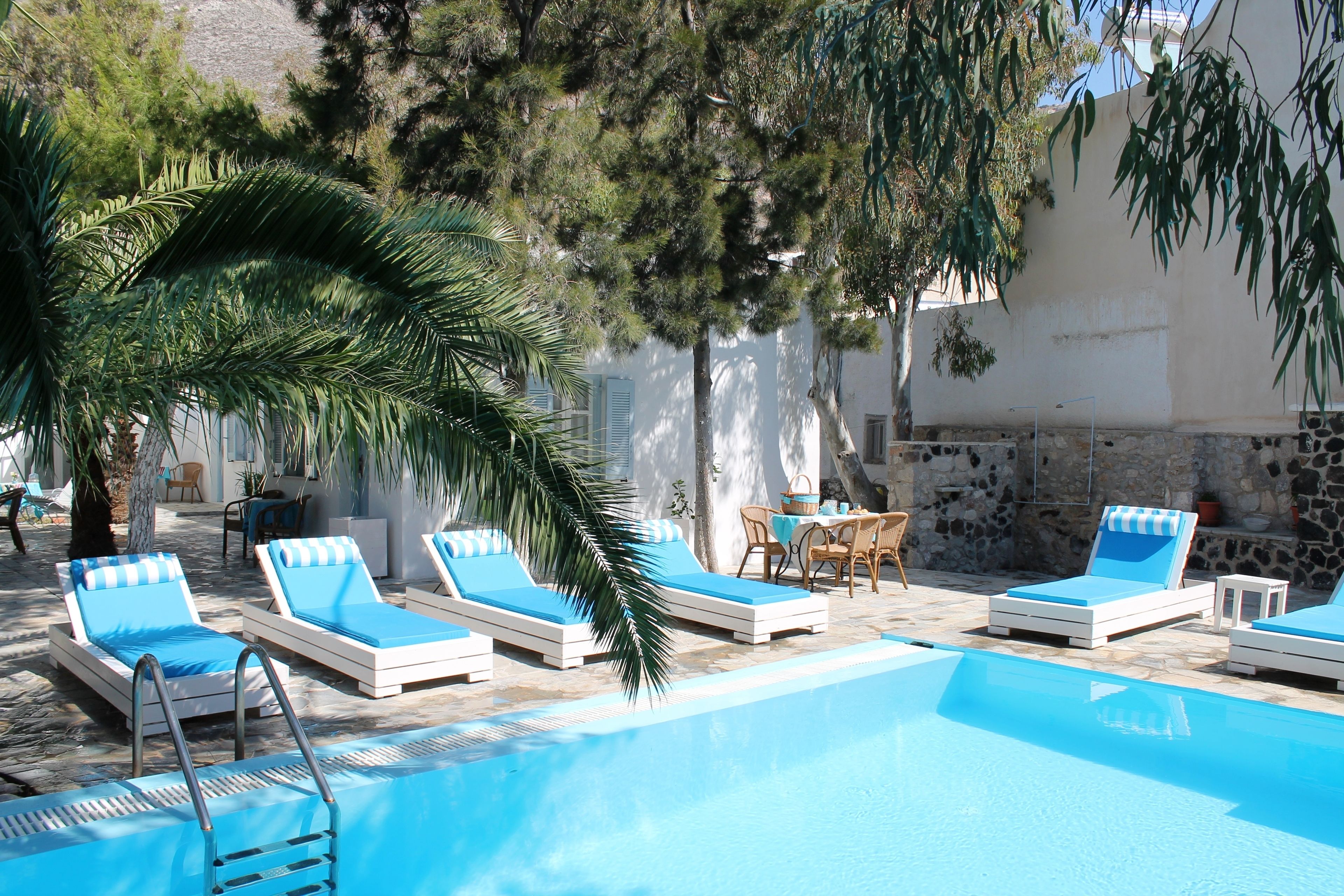 Área junto à piscina com espreguiçadeiras e uma mesa, cercada por árvores e um muro de pedra.