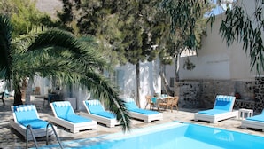 Área junto à piscina com espreguiçadeiras e uma mesa, cercada por árvores e um muro de pedra.