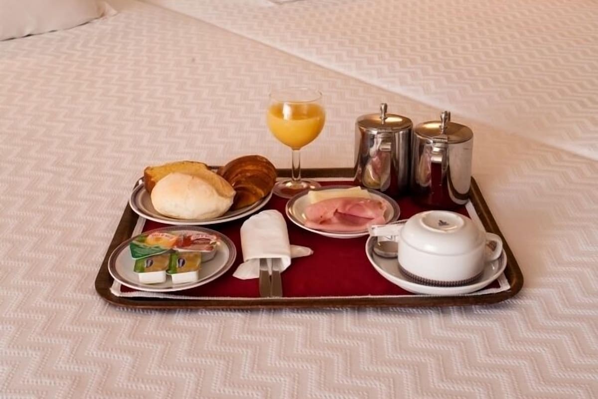 Quarto de hotel com uma cama bem arrumada, mesa de cabeceira com abajur e bandeja de café da manhã com ovos, torradas e suco.