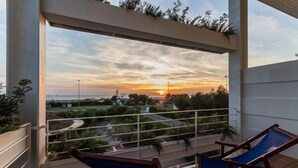 Uma varanda com duas cadeiras de madeira e vista para o pôr do sol sobre uma paisagem com vegetação e edifícios.