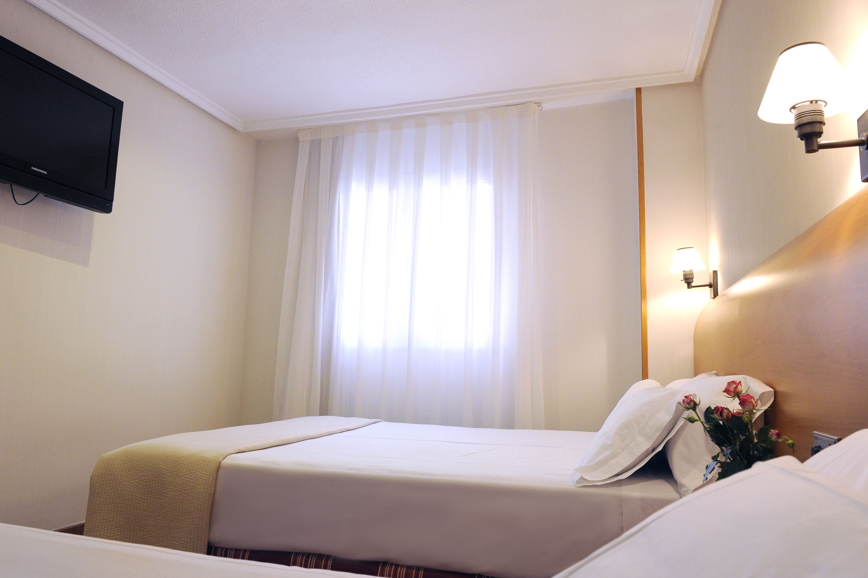 Quarto de hotel com duas camas, uma televisão de tela plana na parede, uma janela com cortinas leves e uma luminária também fixada na parede.