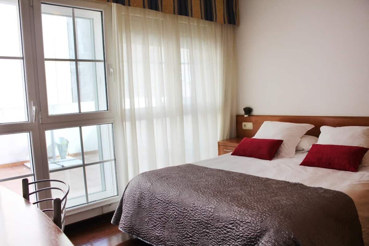 Quarto de hotel com uma janela grande, uma cama com colcha marrom, almofadas brancas e vermelhas, e uma mesa de cabeceira de madeira com uma luminária.