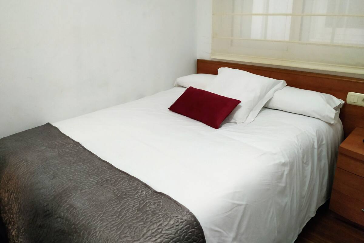 Cama bem arrumada com lençóis brancos e uma almofada vermelha, mesa de cabeceira de madeira com gaveta e janela com persianas.