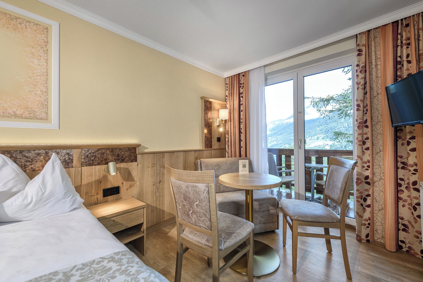 Quarto de hotel com cama, escrivaninha, cadeira, mesa pequena e varanda com vista para as montanhas.