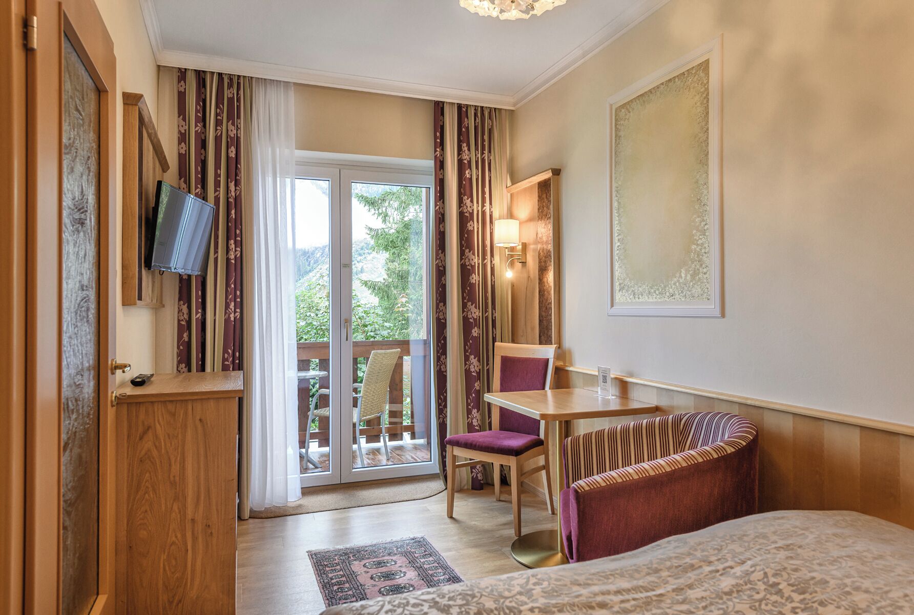 Quarto de hotel com cama, escrivaninha com cadeira, televisão, janela com cortinas e vista para o verde lá fora.