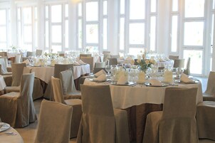 Uma sala de banquetes com mesas redondas preparadas para refeições, grandes janelas e cadeiras com tecido.