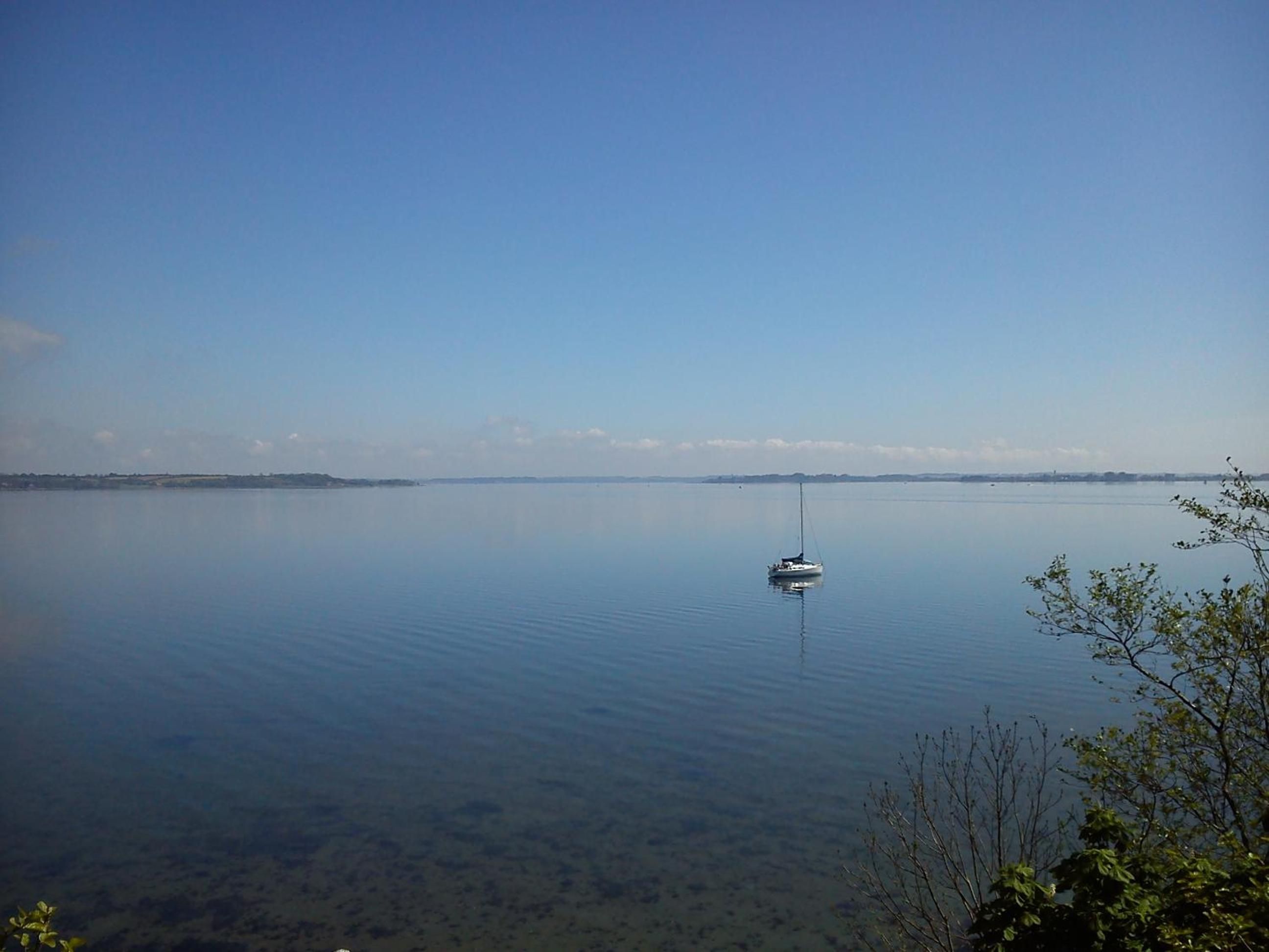 Um lago tranquilo com um veleiro ancorado no centro, rodeado por águas cristalinas e terra distante.