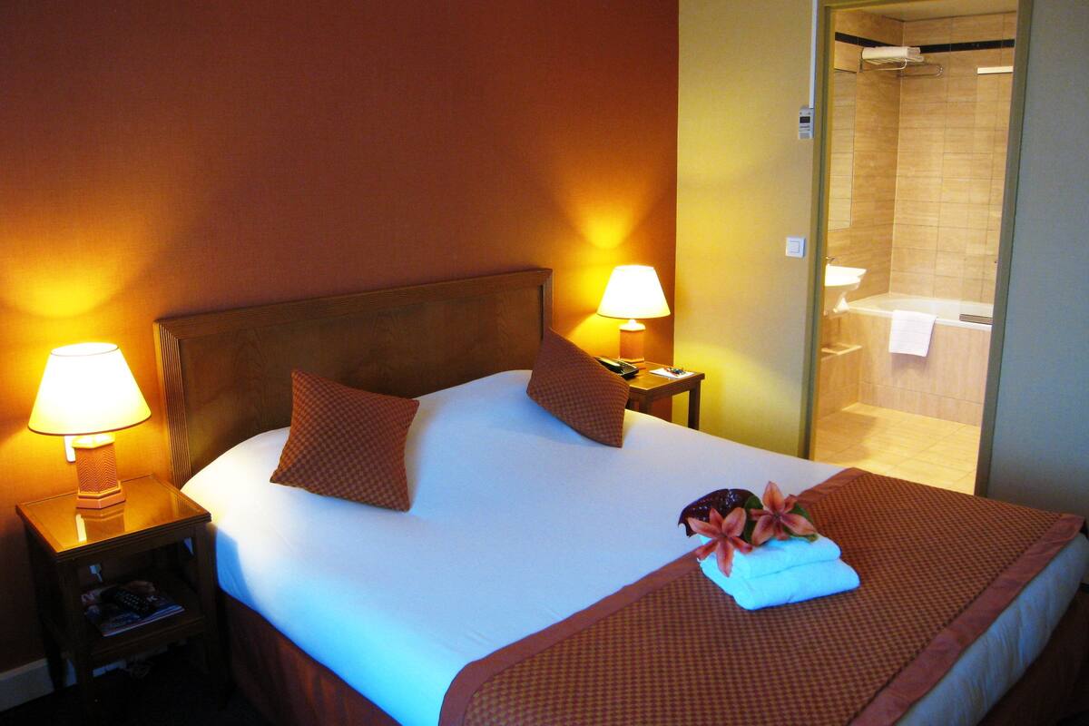 Quarto de hotel com uma cama grande, duas luminárias de cabeceira, mesa de cabeceira e vista para um banheiro com banheira e chuveiro.