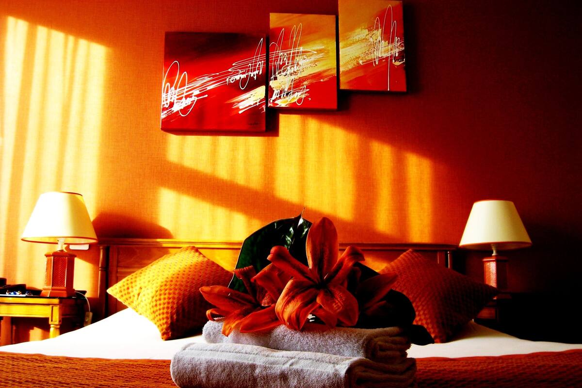 Quarto de hotel com uma cama, duas luminárias de cabeceira, um arranjo floral decorativo e três obras de arte nas paredes.