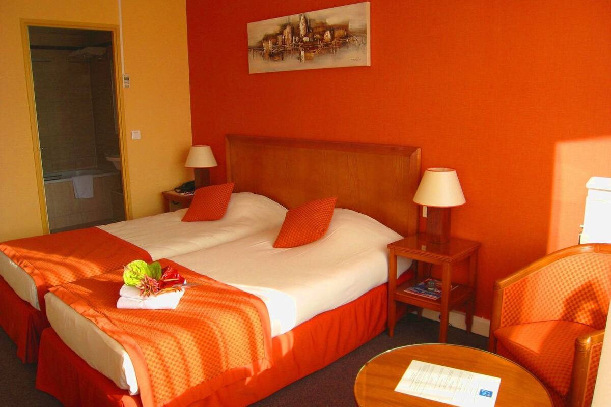 Quarto de hotel com uma cama grande, duas almofadas, mesa de cabeceira com luminária, uma cadeira, uma pequena mesa e um quadro na parede.