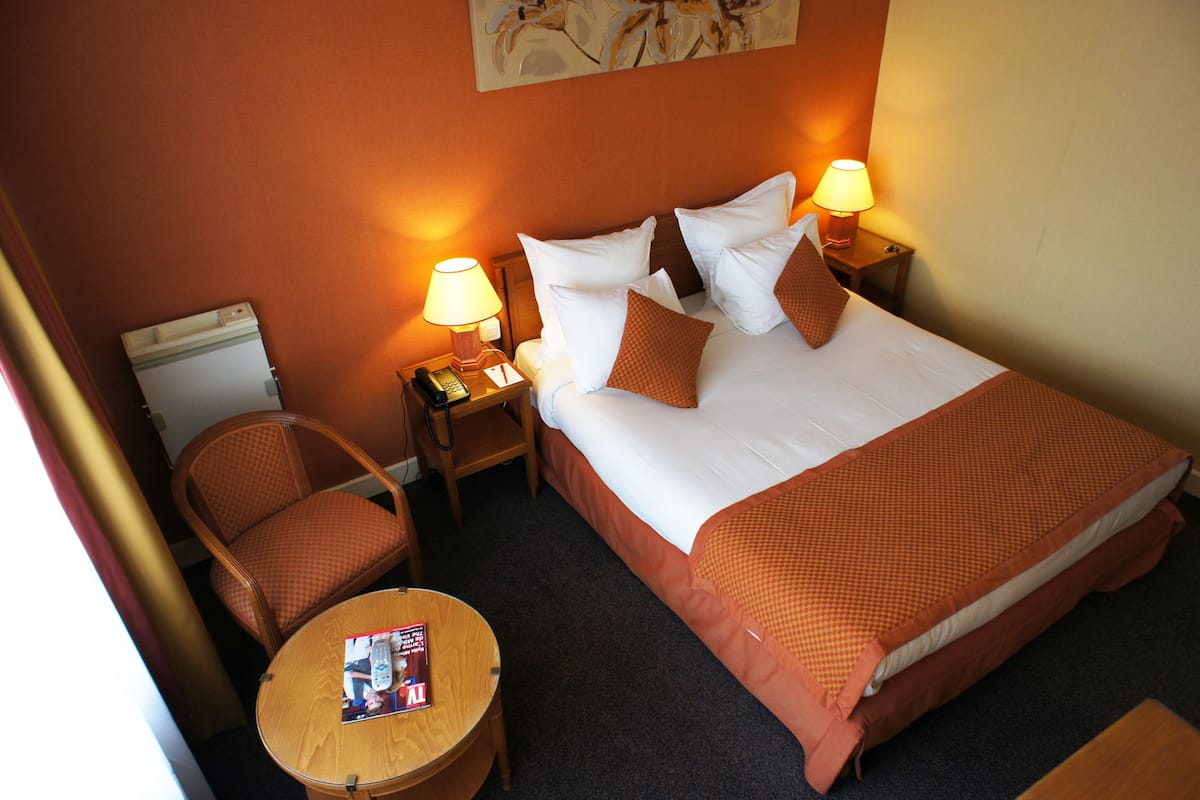 Quarto de hotel com uma cama, duas luminárias de cabeceira, uma cadeira, uma mesinha de centro com uma revista e um aquecedor.