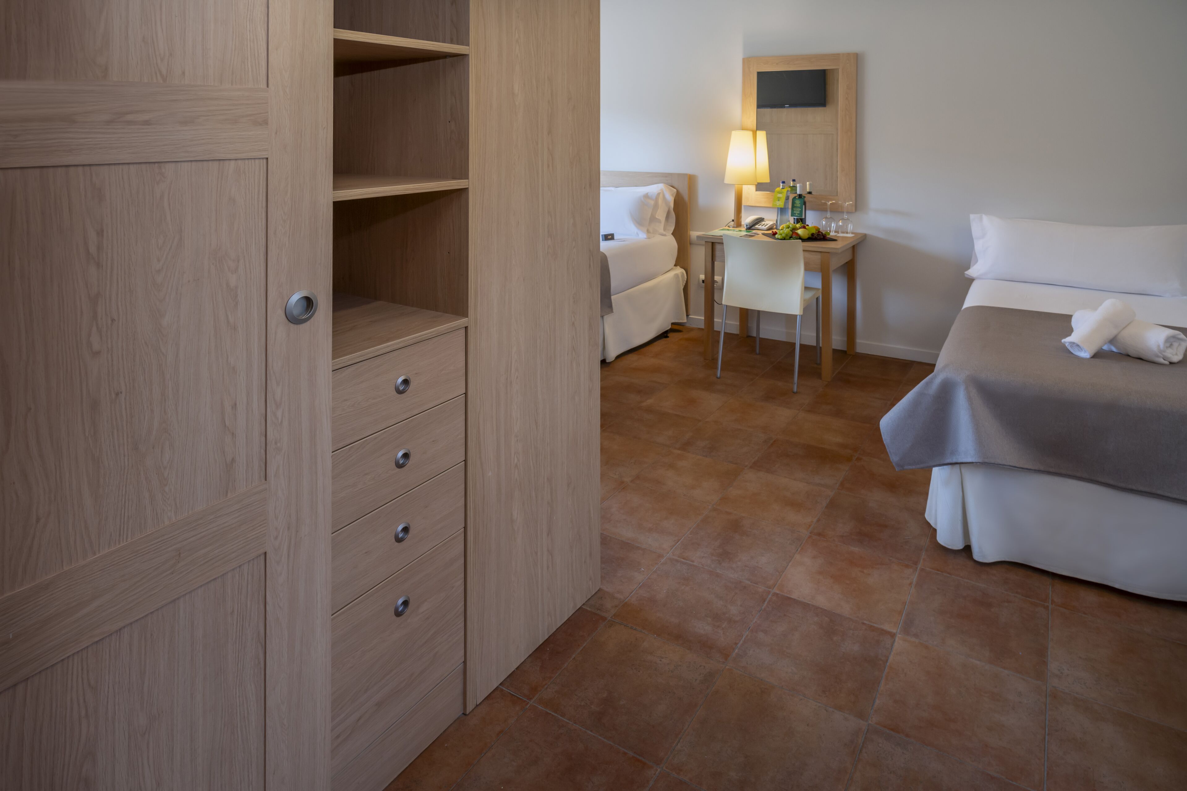 Quarto de hotel com guarda-roupa de madeira, cama com colcha cinzenta, secretária pequena com cadeira e uma luminária.