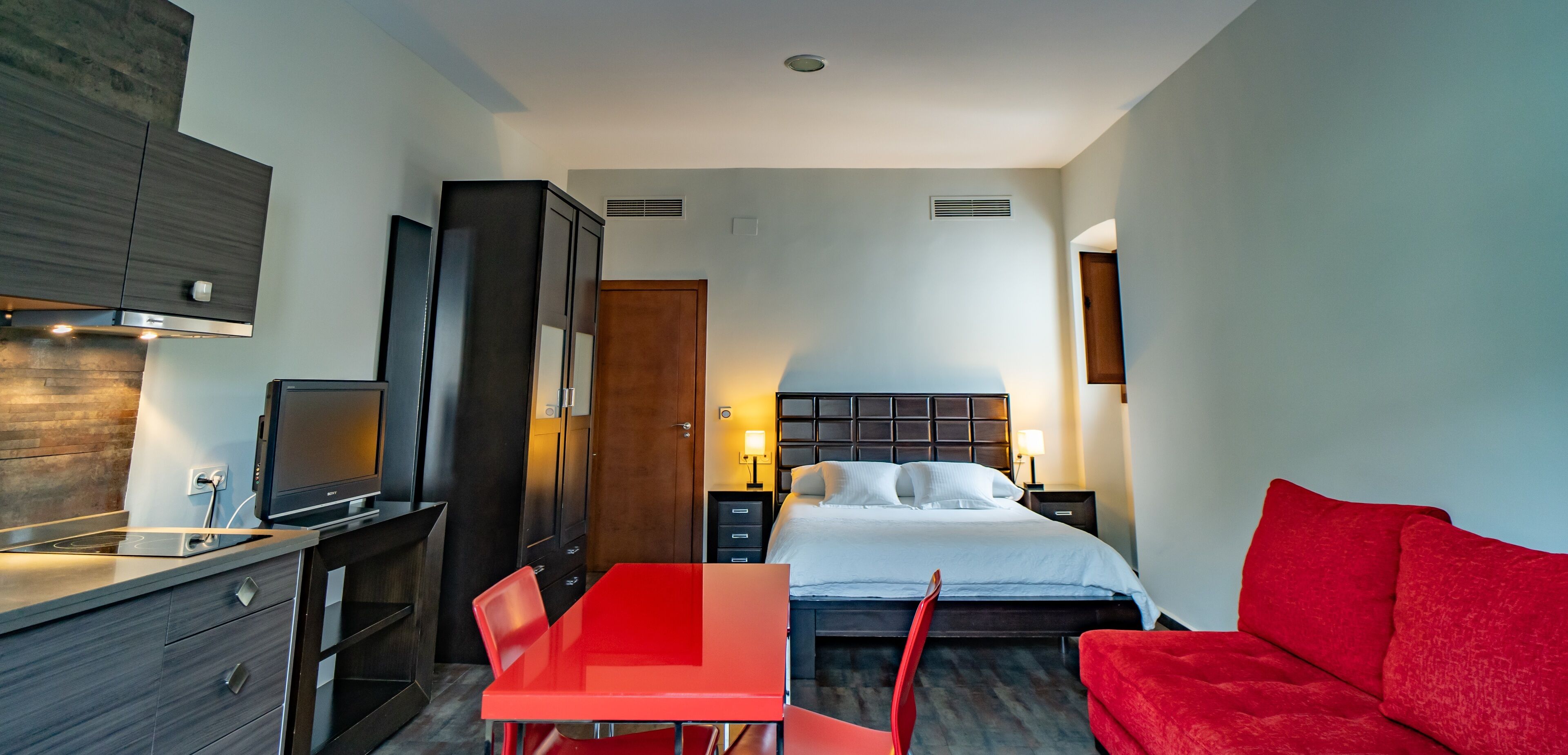 Quarto de hotel compacto com cama, sofá vermelho, escrivaninha com cadeira, televisão e cozinha compacta.
