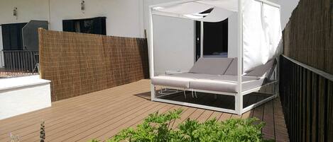 Uma cama branca ao ar livre com dossel, cercada por um deck de madeira e uma cerca de bambu.