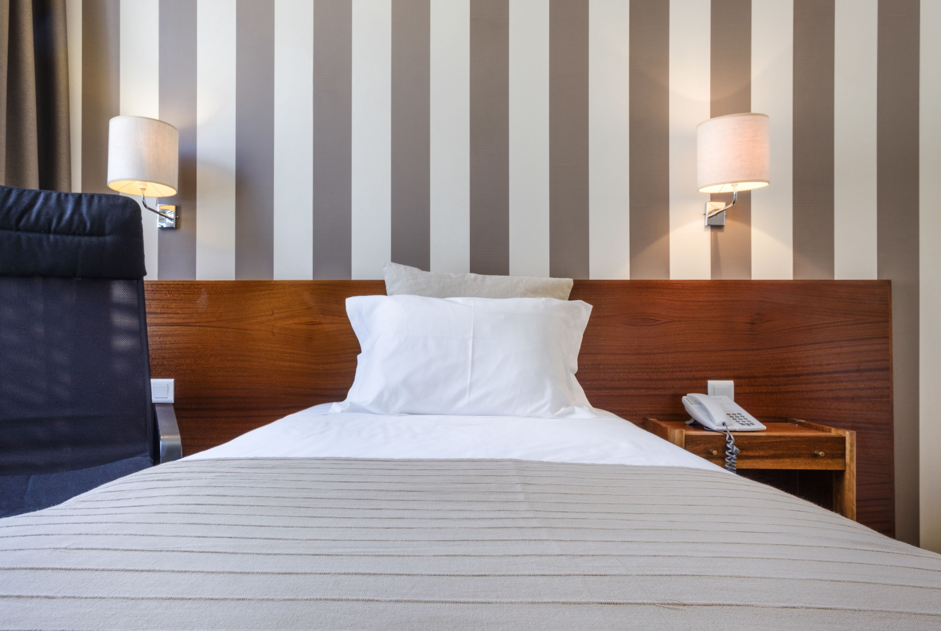 Quarto de hotel com cabeceira de madeira, cama com lençóis brancos, duas luminárias fixadas na parede e mesa de cabeceira com telefone.