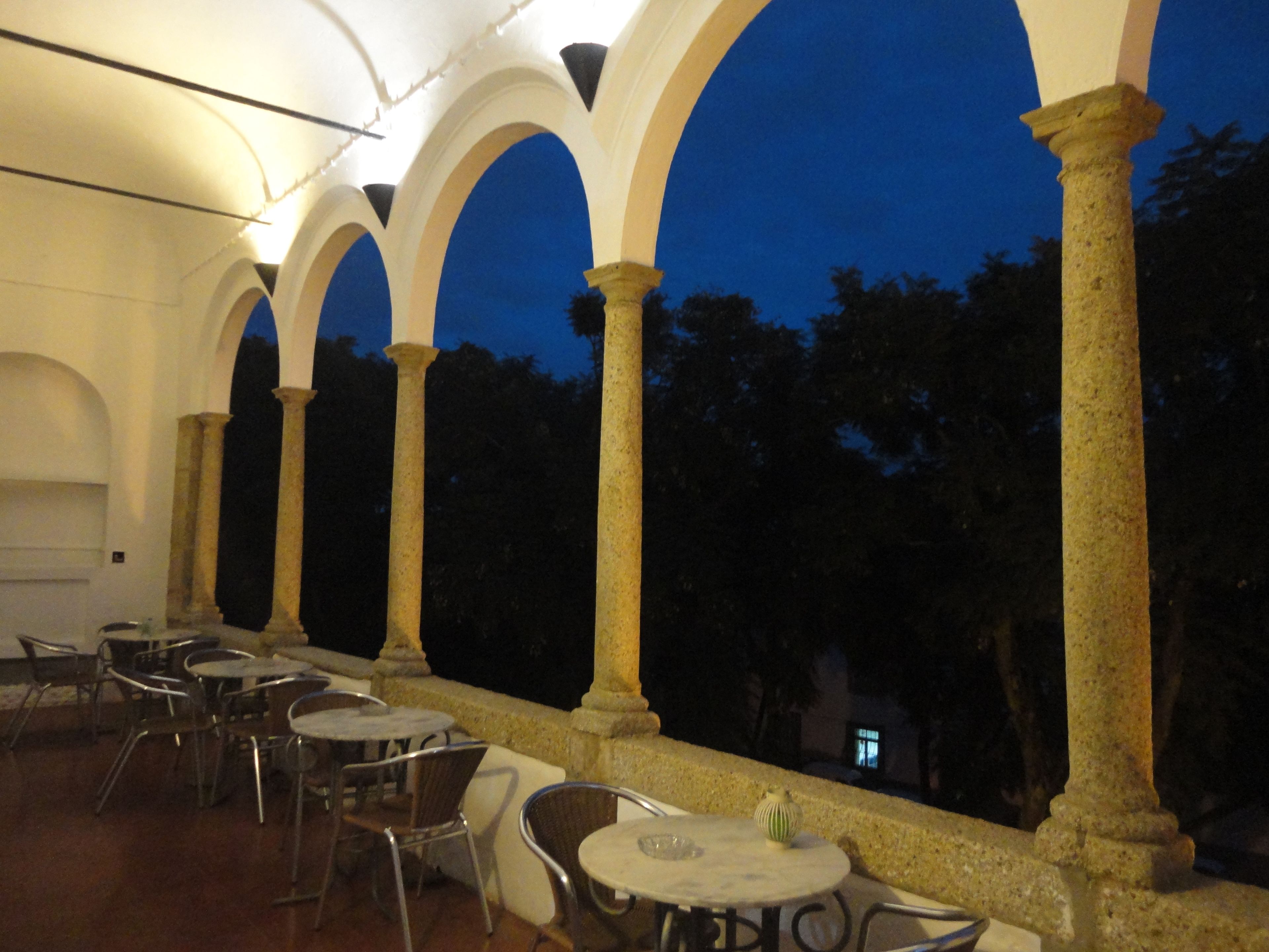 Um pátio externo com mesas e cadeiras redondas, rodeado por colunas e arcos de pedra, sob um céu noturno.