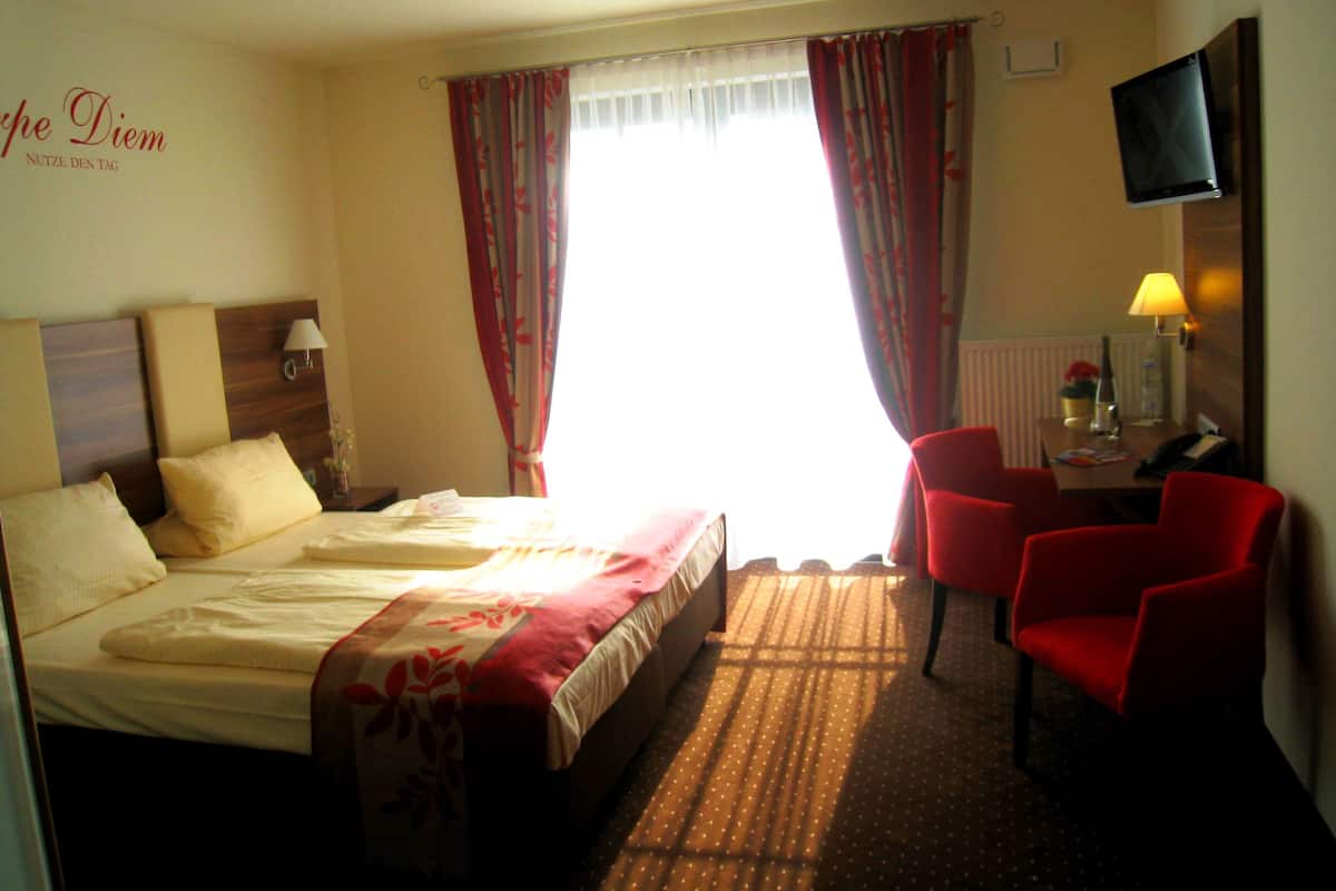 Quarto de hotel com uma cama, duas cadeiras, uma mesa com luminária e uma televisão na parede.