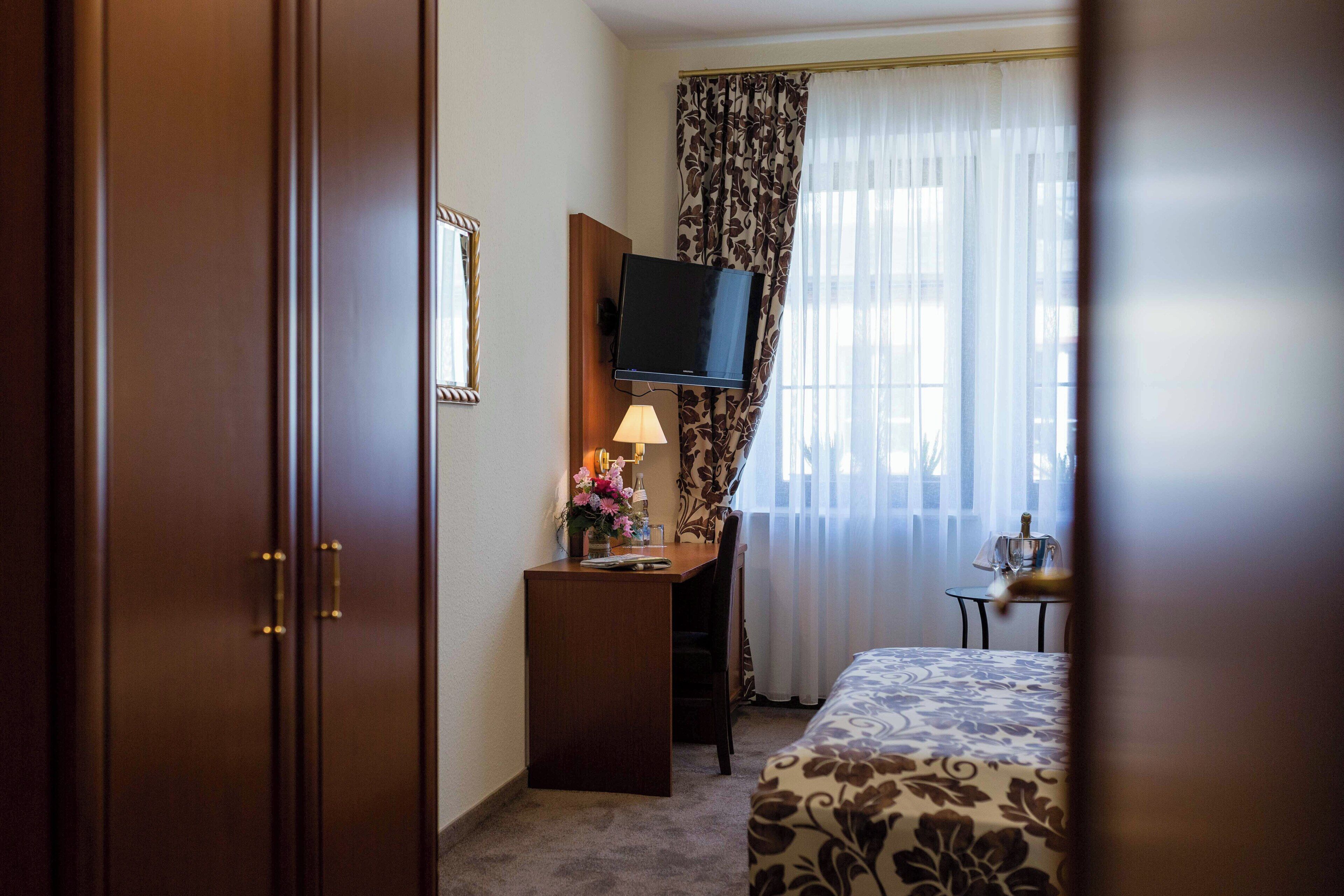 Quarto de hotel com cama, escrivaninha com luminária, televisão e janela com cortinas.