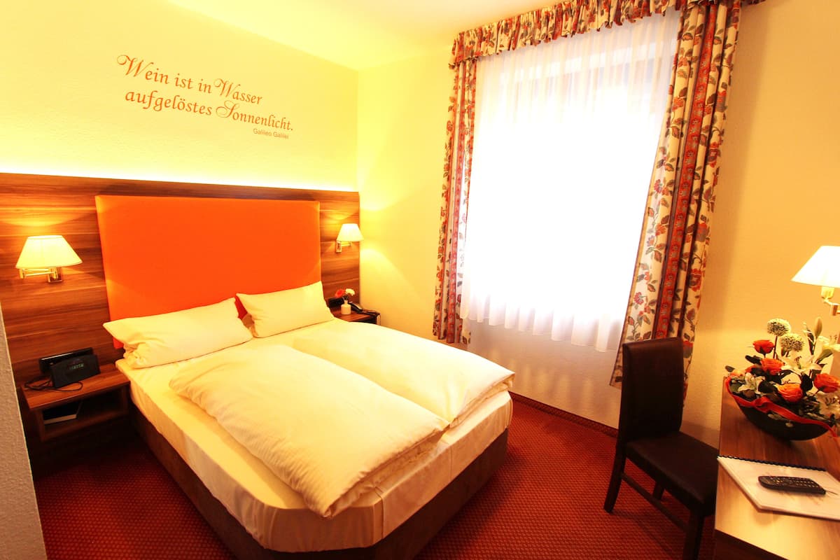 Quarto de hotel com uma cama grande, duas mesas de cabeceira com luminárias, uma cadeira, uma escrivaninha com um controle remoto e um arranjo floral.
