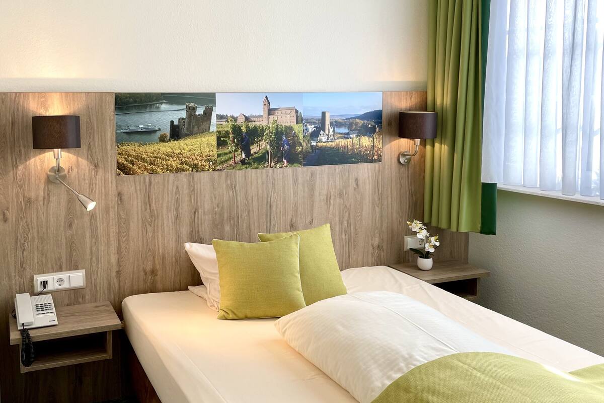 Quarto de hotel com uma cama, duas mesas de cabeceira, uma luminária, um telefone e um quadro na parede retratando uma paisagem.