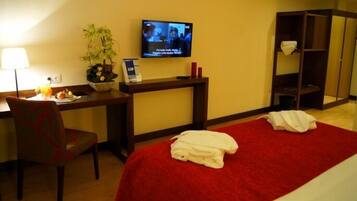 Quarto de hotel com cama, secretária com computador, televisão, planta e tapete vermelho.
