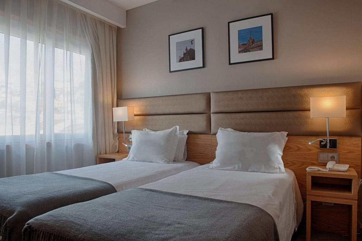 Quarto de hotel com duas camas, uma janela grande com cortinas translúcidas, duas quadros na parede e luminárias de cabeceira.