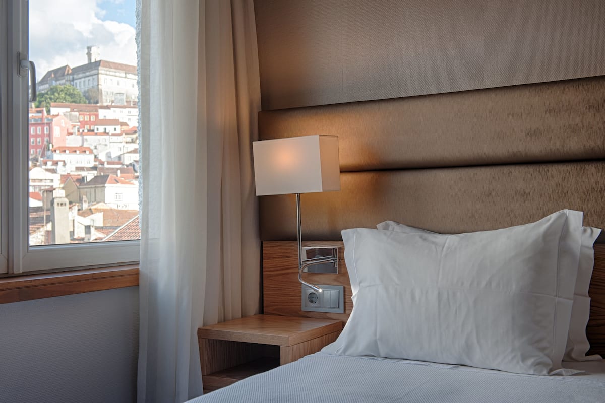 Quarto de hotel com cama, mesa de cabeceira, abajur e vista para a cidade pela janela.