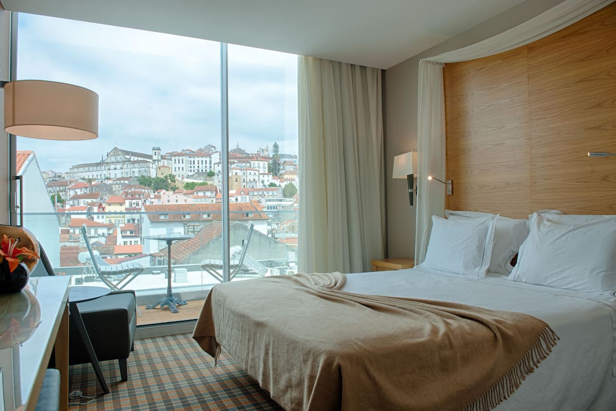 Quarto de hotel com uma cama grande, uma escrivaninha com abajur e vista para a paisagem urbana através de grandes janelas.