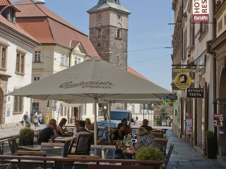 Um café ao ar livre com mesas e cadeiras, um letreiro indicando restaurante de hotel, e um edifício com uma torre ao fundo.