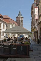 Um café ao ar livre com mesas e cadeiras, um letreiro indicando restaurante de hotel, e um edifício com uma torre ao fundo.