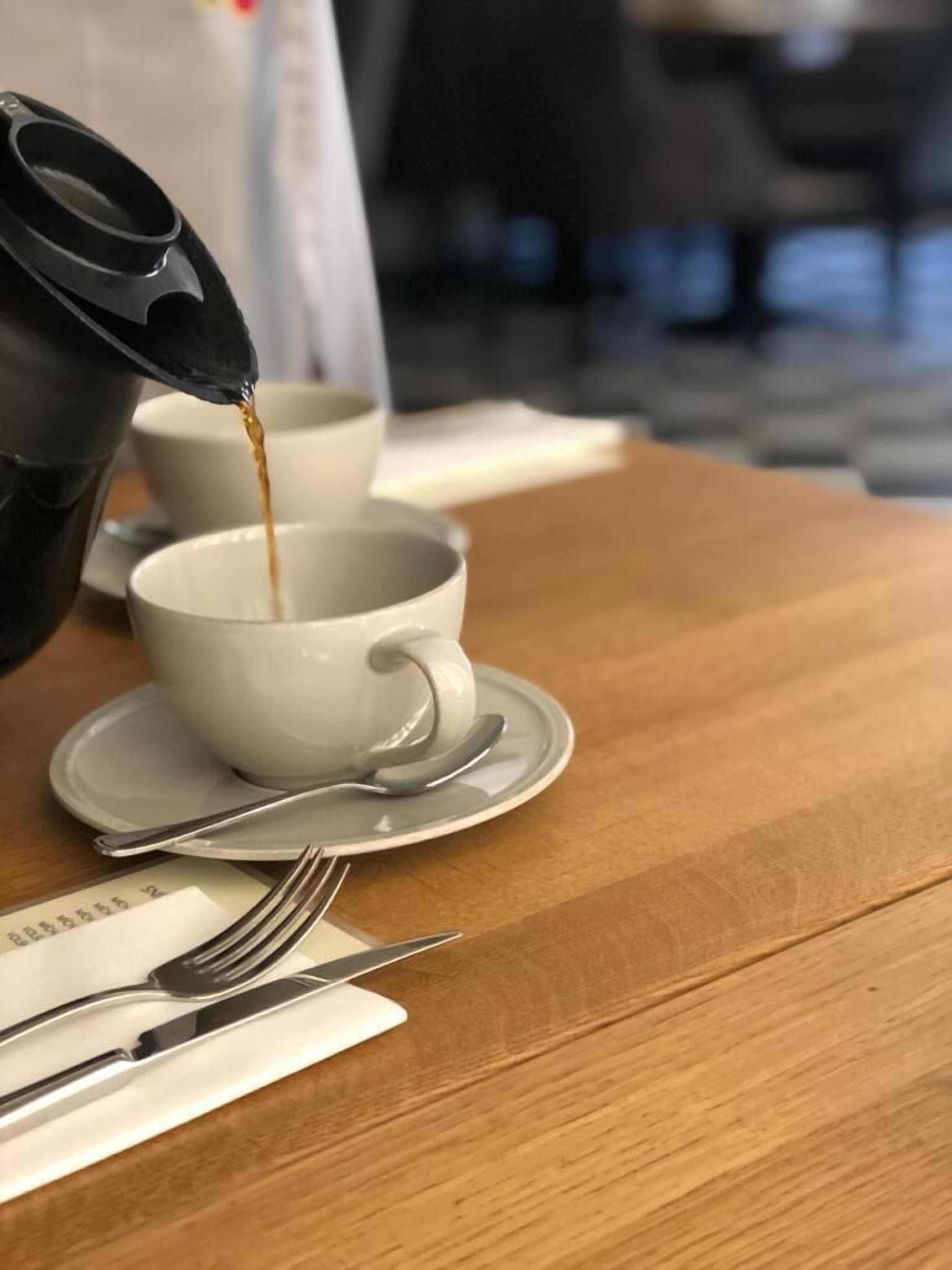 Uma cafeteira servindo café numa chávena branca sobre uma mesa de madeira.