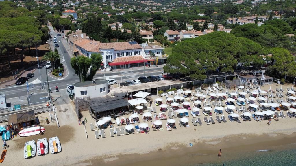 Vista aérea de uma praia com fileiras de espreguiçadeiras e guarda-sóis, uma estrada com carros e edifícios ao fundo.