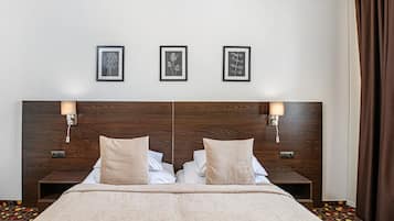 Uma cama de casal com cabeceira de madeira, duas mesas de cabeceira com luminárias e três impressões botânicas emolduradas na parede.
