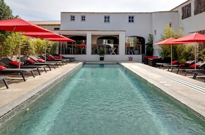 Piscina com espreguiçadeiras e guarda-sóis vermelhos, cercada por um edifício com várias janelas e telhado em azulejos.