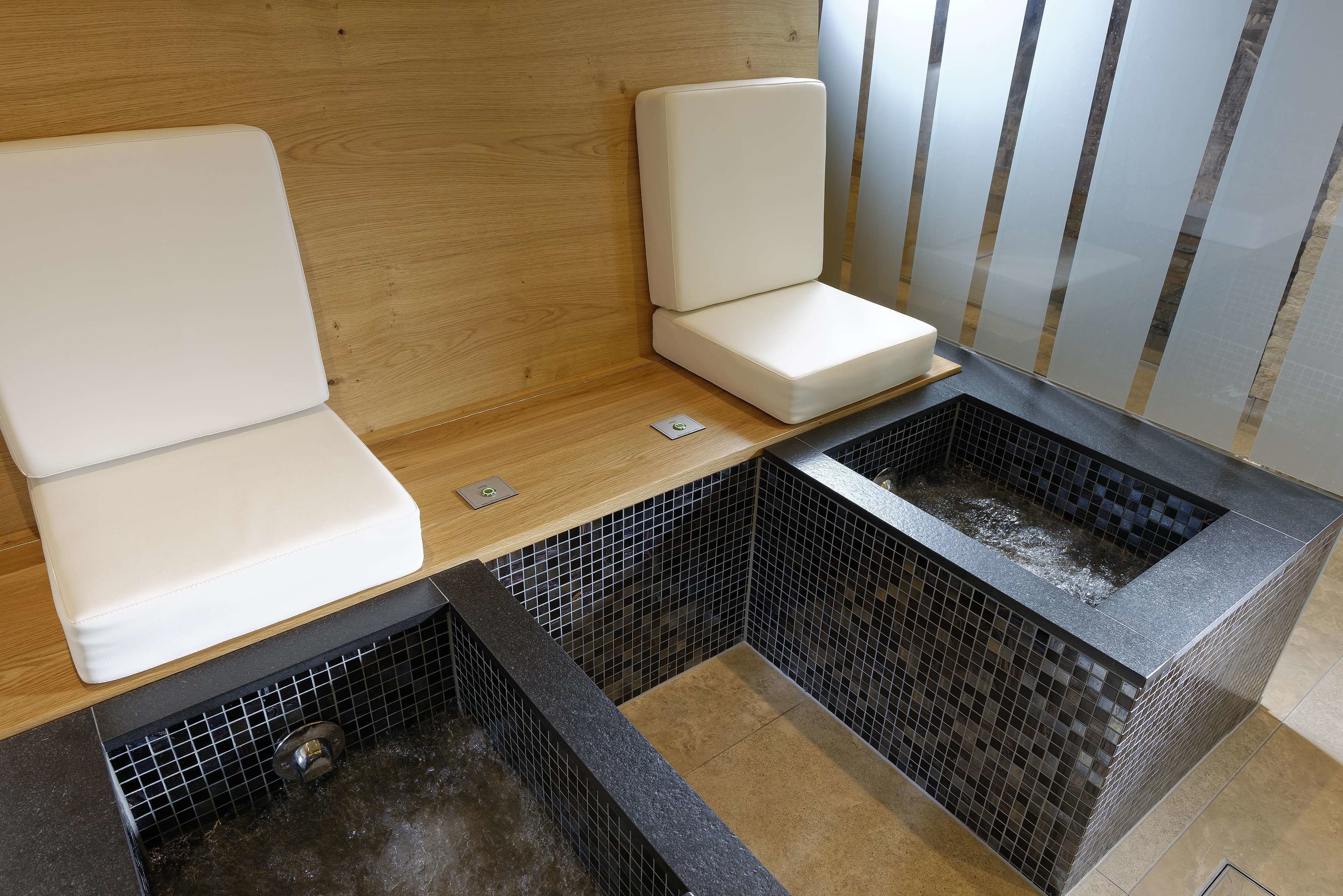 Uma área de spa com dois bancos brancos acolchoados, piso ladrilhado e um pequeno elemento de água.