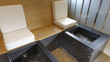 Uma área de spa com dois bancos brancos acolchoados, piso ladrilhado e um pequeno elemento de água.