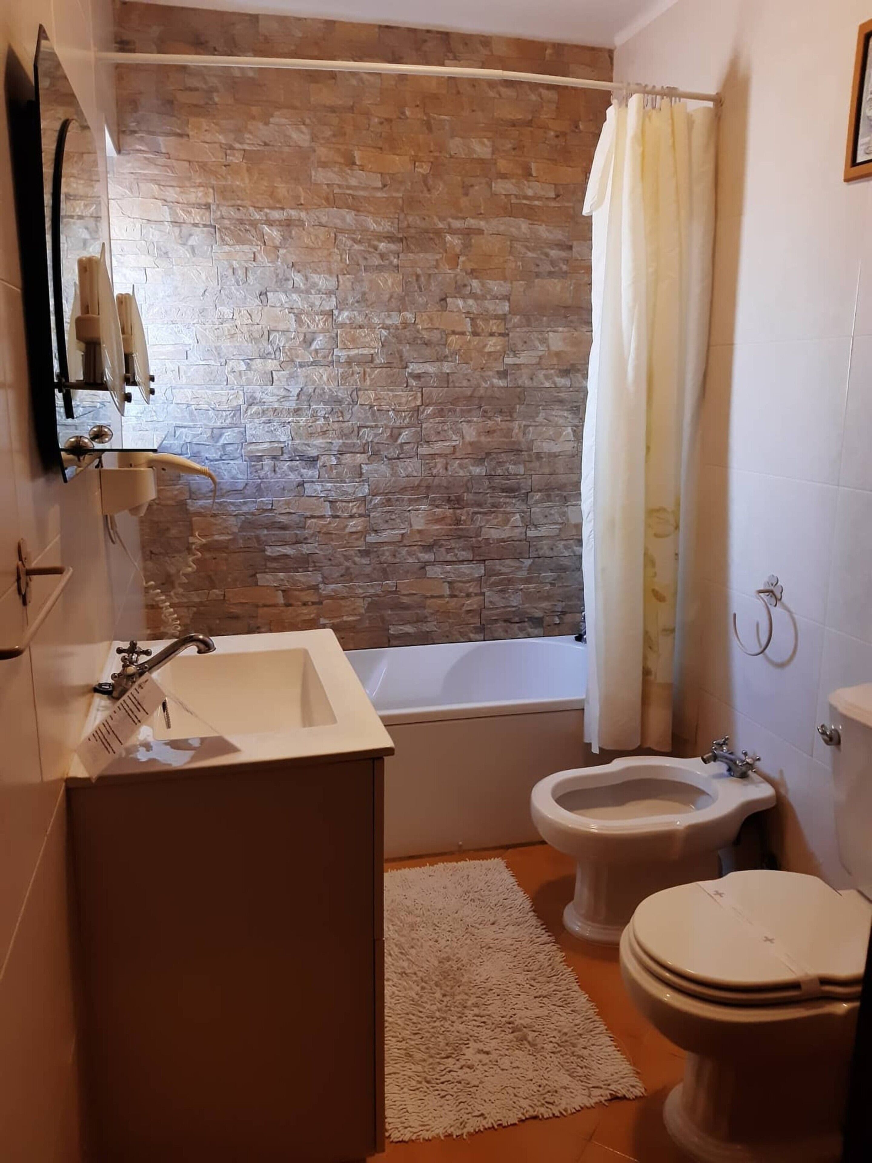 Casa de banho com parede de pedra, banheira, bidé, sanita e lavatório com espelho em cima.