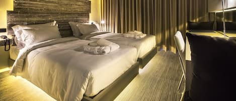Quarto de hotel moderno com uma cama grande, uma mesinha de cabeceira, um abajur e vista para a cidade à noite.