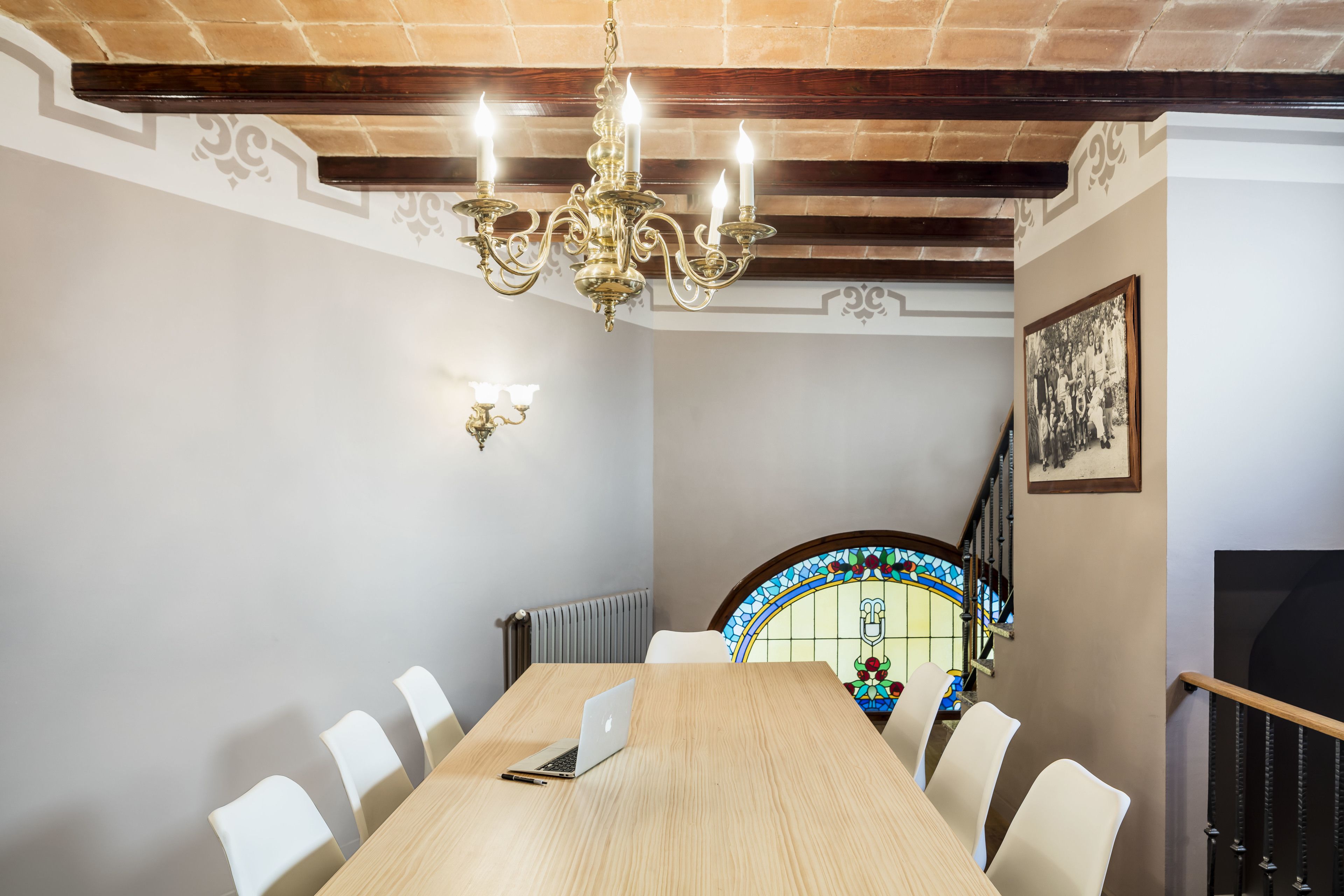 Uma sala de conferências com mesa de madeira, cadeiras brancas, um candelabro e uma janela de vitrais.