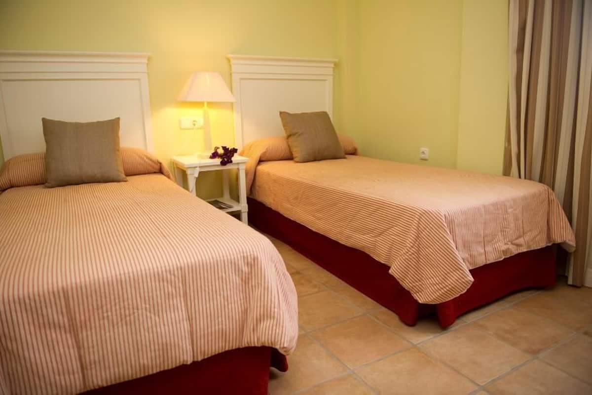Quarto de hotel com duas camas, cada uma com colcha e almofadas listradas, uma mesa de cabeceira com abajur e uma janela com cortinas.