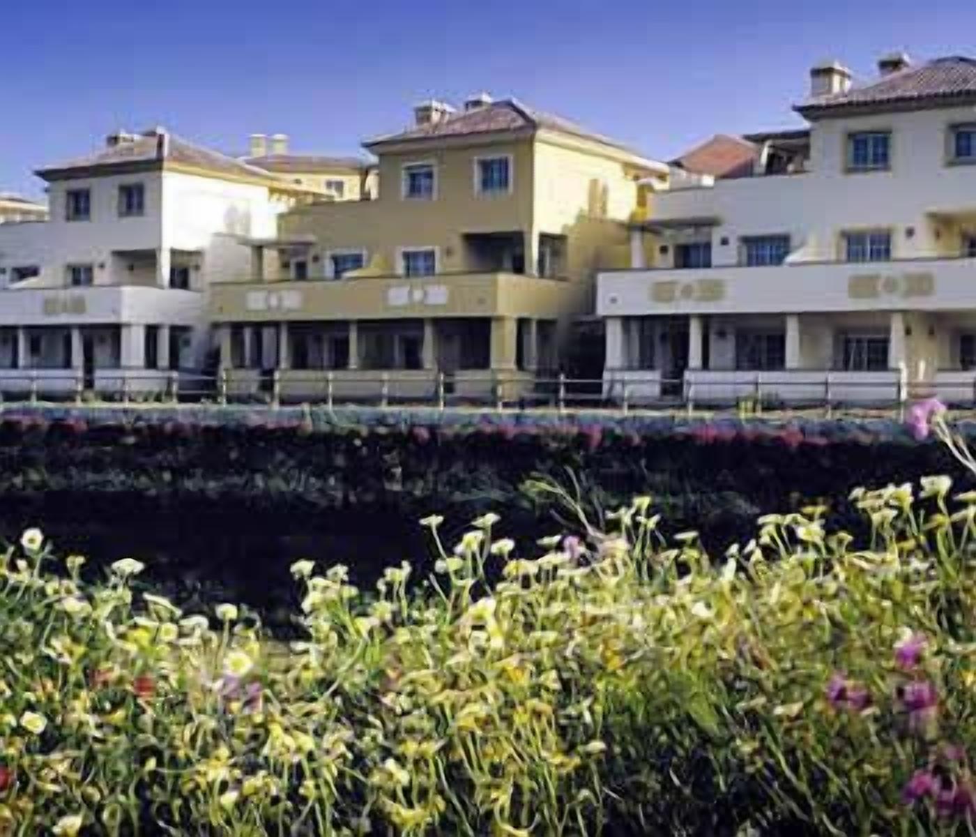 Uma fileira de edifícios residenciais modernos, com dois andares e varandas, localizada perto de um corpo d'água, com flores em plena floração ao primeiro plano.
